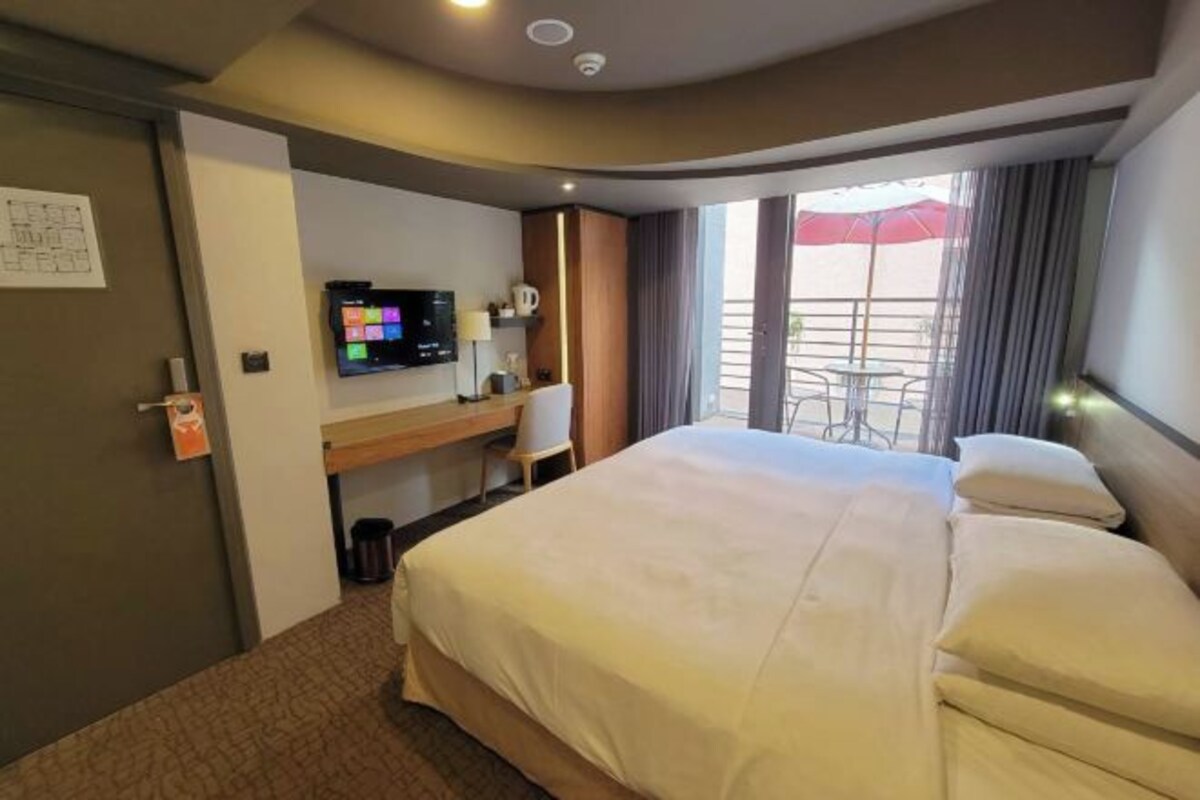 Quarto de hotel com uma cama grande, uma escrivaninha com televisão de tela plana, uma cadeira e uma varanda com mesa e guarda-sol.
