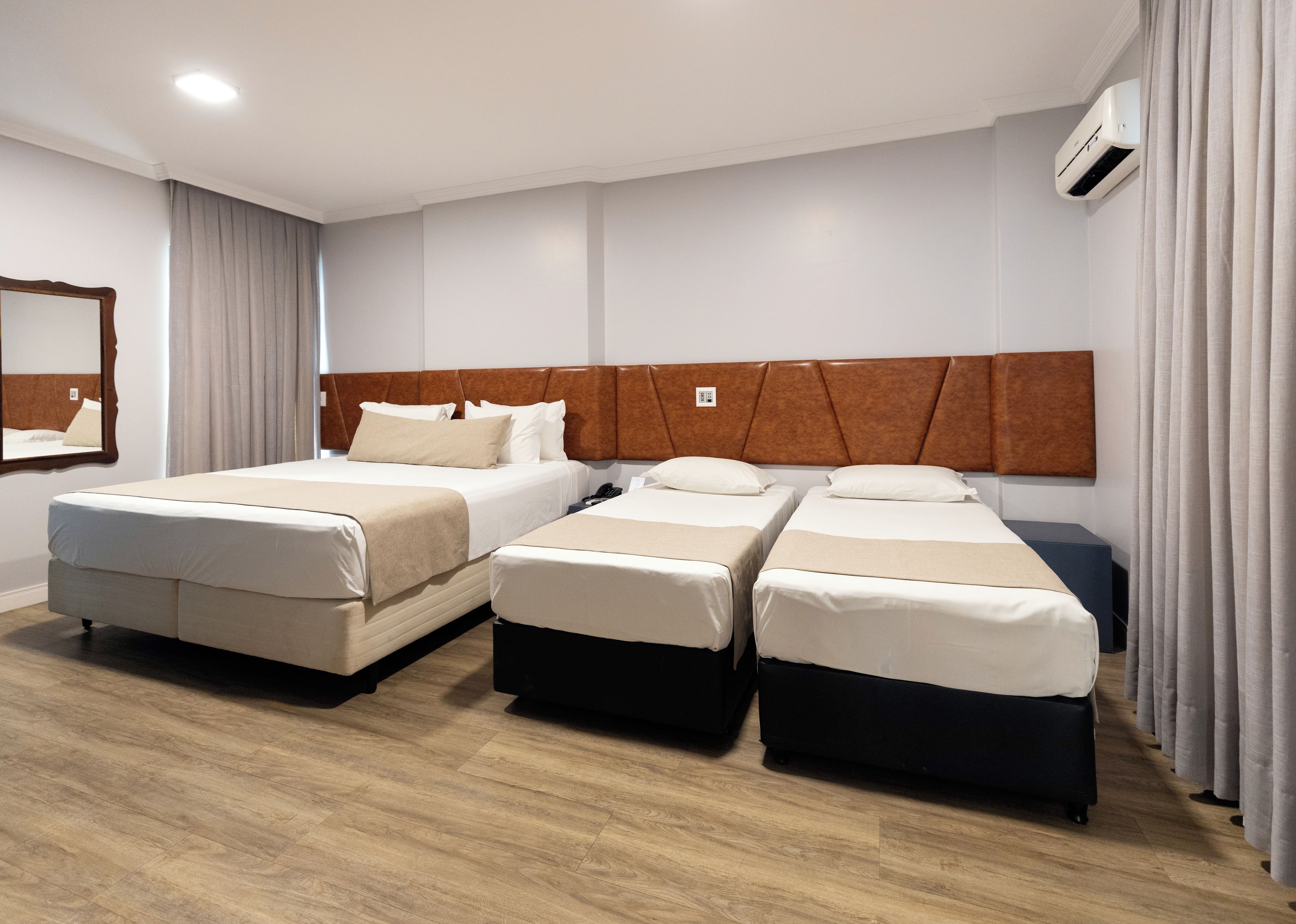 Quarto de hotel com três camas, cabeceira de madeira, espelho e ar condicionado.