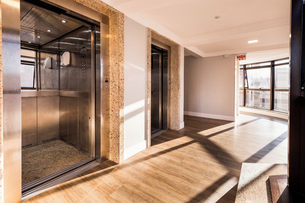 Um elevador moderno com interior metálico, próximo a um corredor com soalho de madeira e grandes janelas.