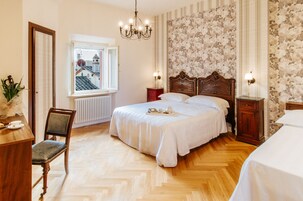 Quarto com uma cama grande, uma cadeira de madeira, uma mesa pequena com um vaso e uma janela com vista para prédios.