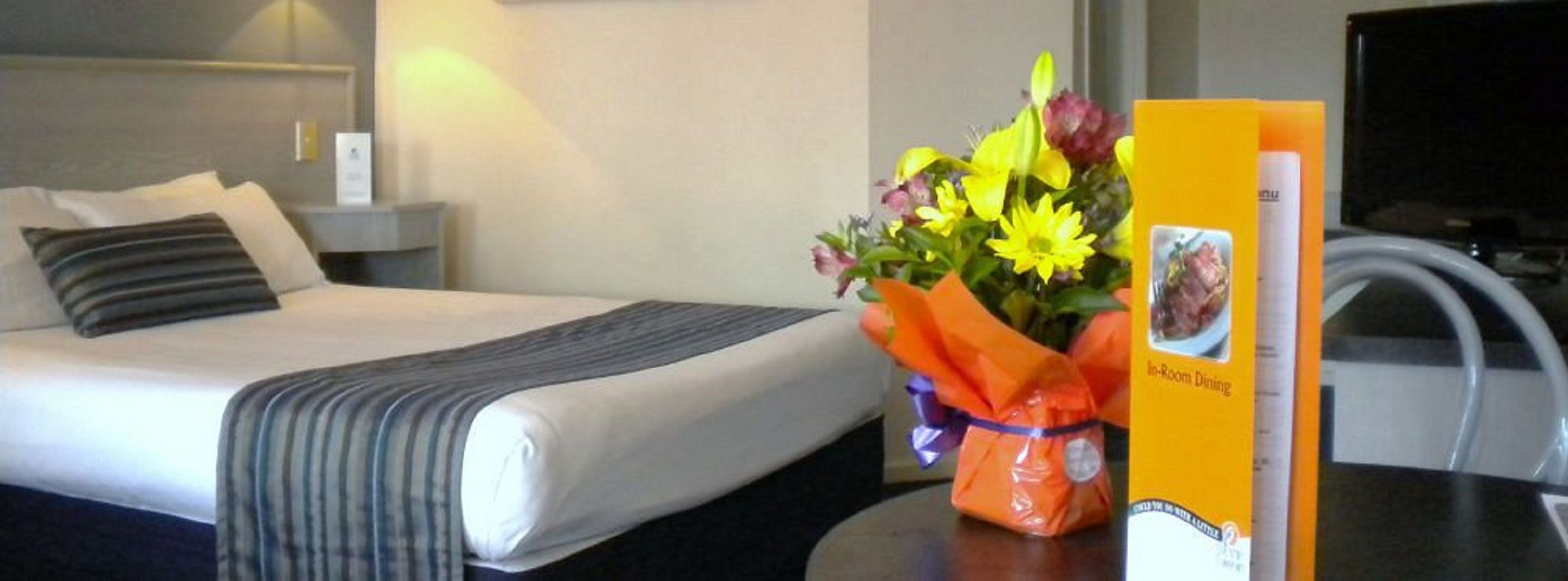 Quarto de hotel com uma cama bem arrumada, mesa de cabeceira com um arranjo de flores e um folheto sobre a mesa.