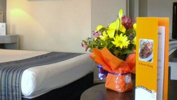 Quarto de hotel com uma cama bem arrumada, mesa de cabeceira com um arranjo de flores e um folheto sobre a mesa.