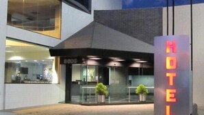 Entrada de hotel com letreiro de neon, portas de vidro e plantas em vasos.