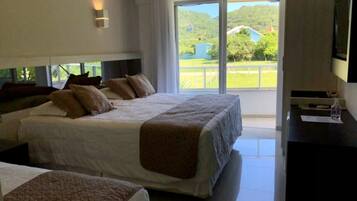 Quarto de hotel com duas camas, televisão, escrivaninha e varanda com vista para áreas verdes e edifícios.