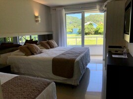 Quarto de hotel com duas camas, televisão, escrivaninha e varanda com vista para áreas verdes e edifícios.
