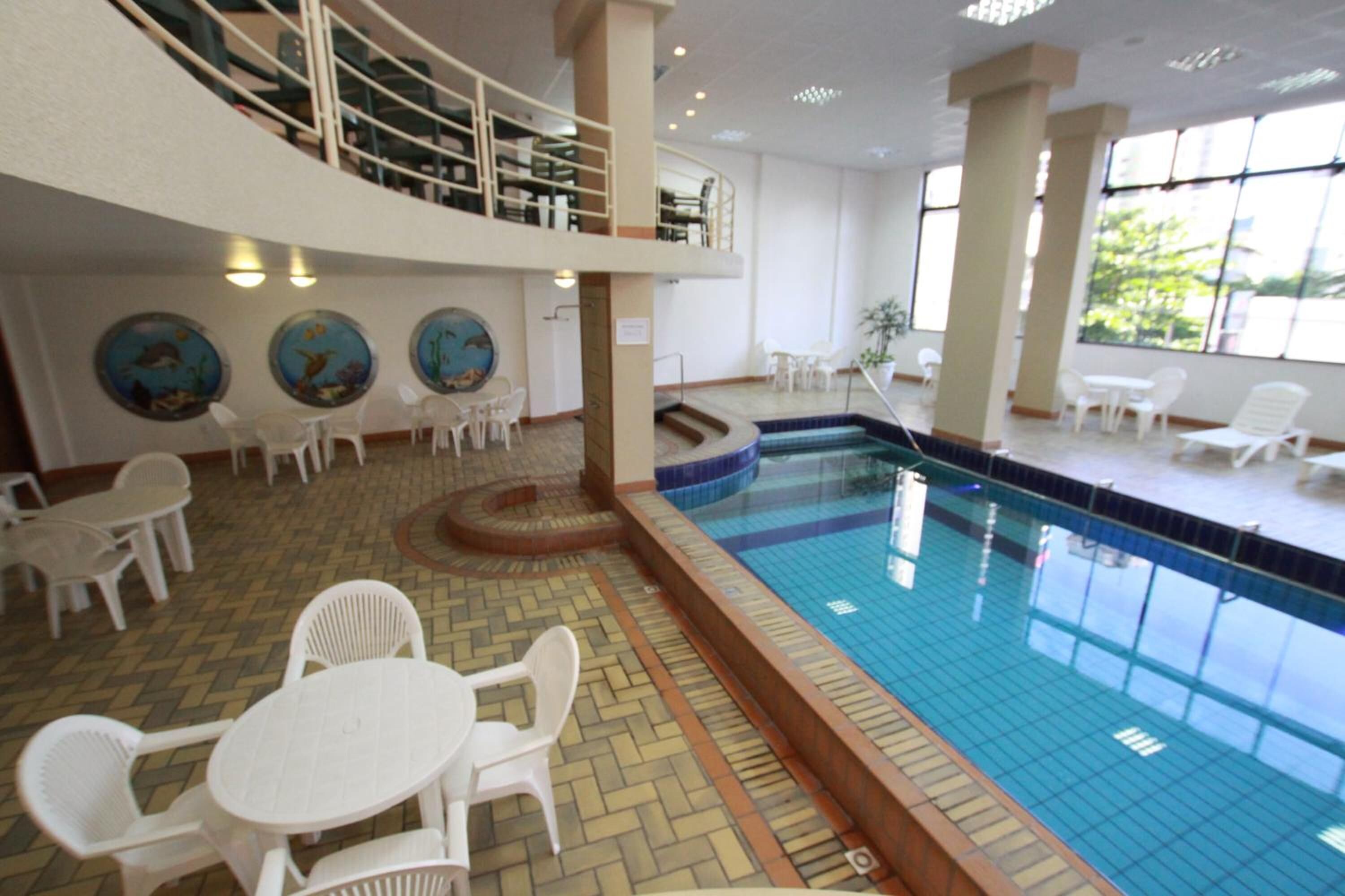 Piscina interna com chão de azulejos, cercada por cadeiras e mesas de plástico branco.