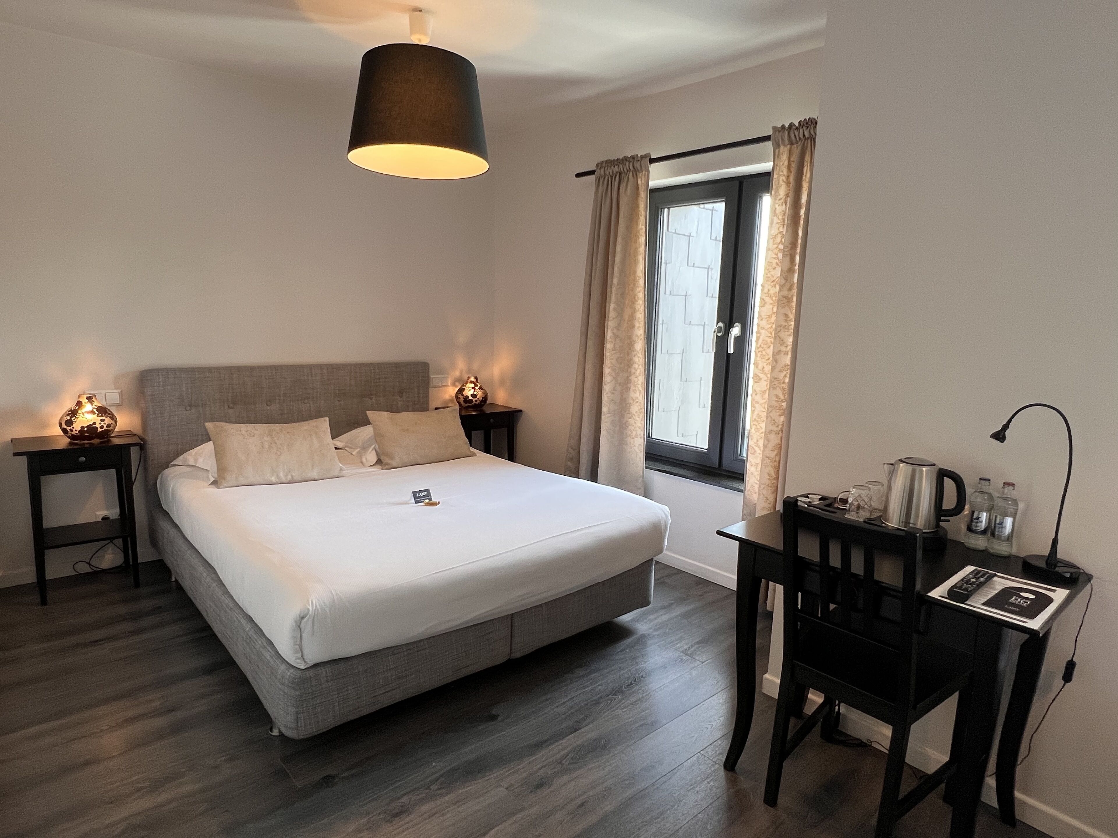 Um quarto com cama, mesas de cabeceira, escrivaninha com cadeira, janela com cortinas e luminária pendente.