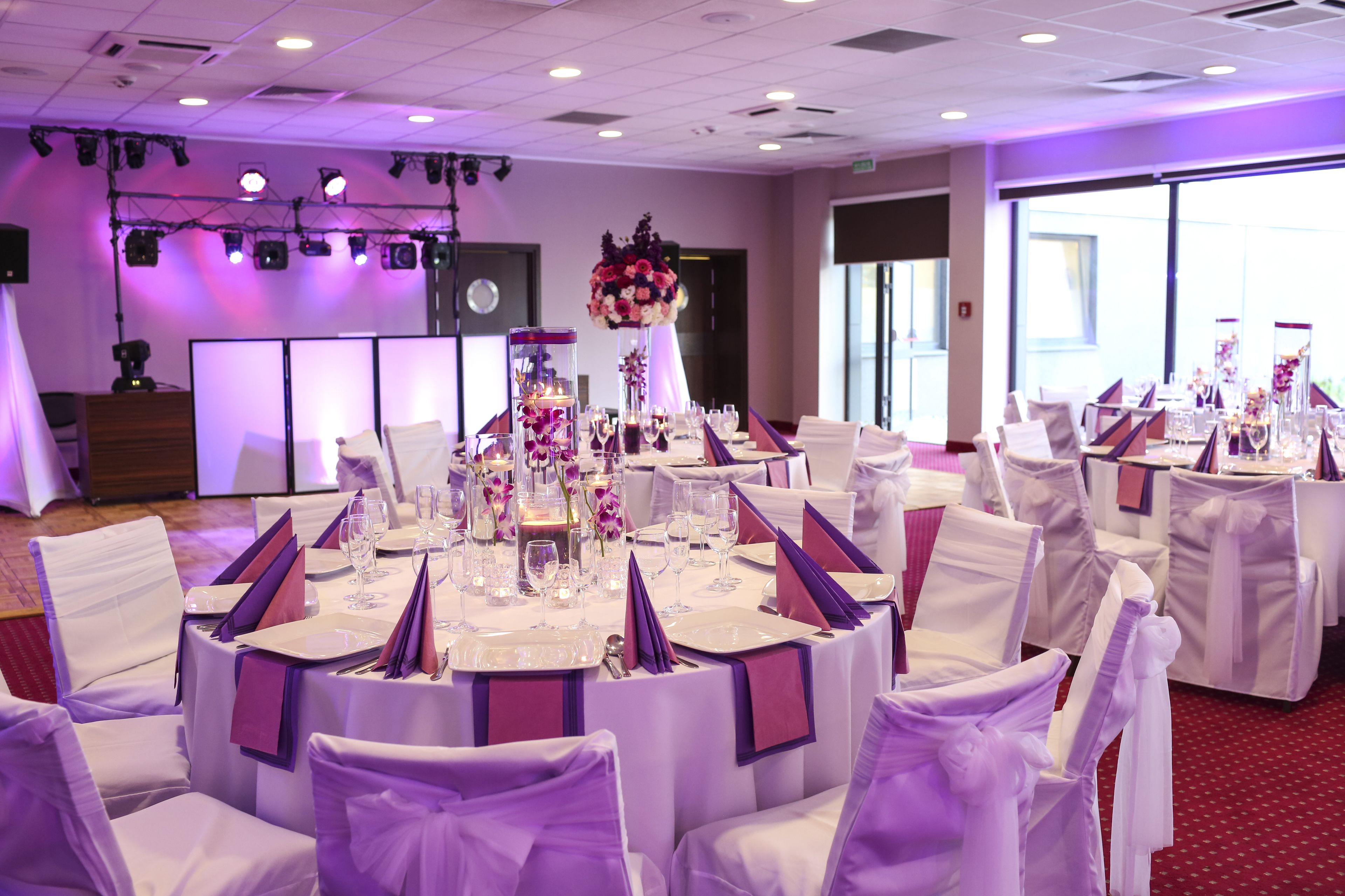 Uma sala de banquetes com mesas redondas preparadas para um evento formal, com toalhas brancas e guardanapos roxos.