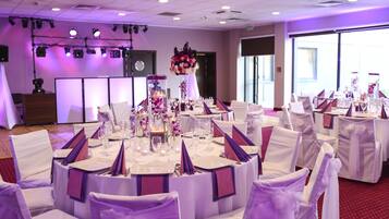 Uma sala de banquetes com mesas redondas preparadas para um evento formal, com toalhas brancas e guardanapos roxos.
