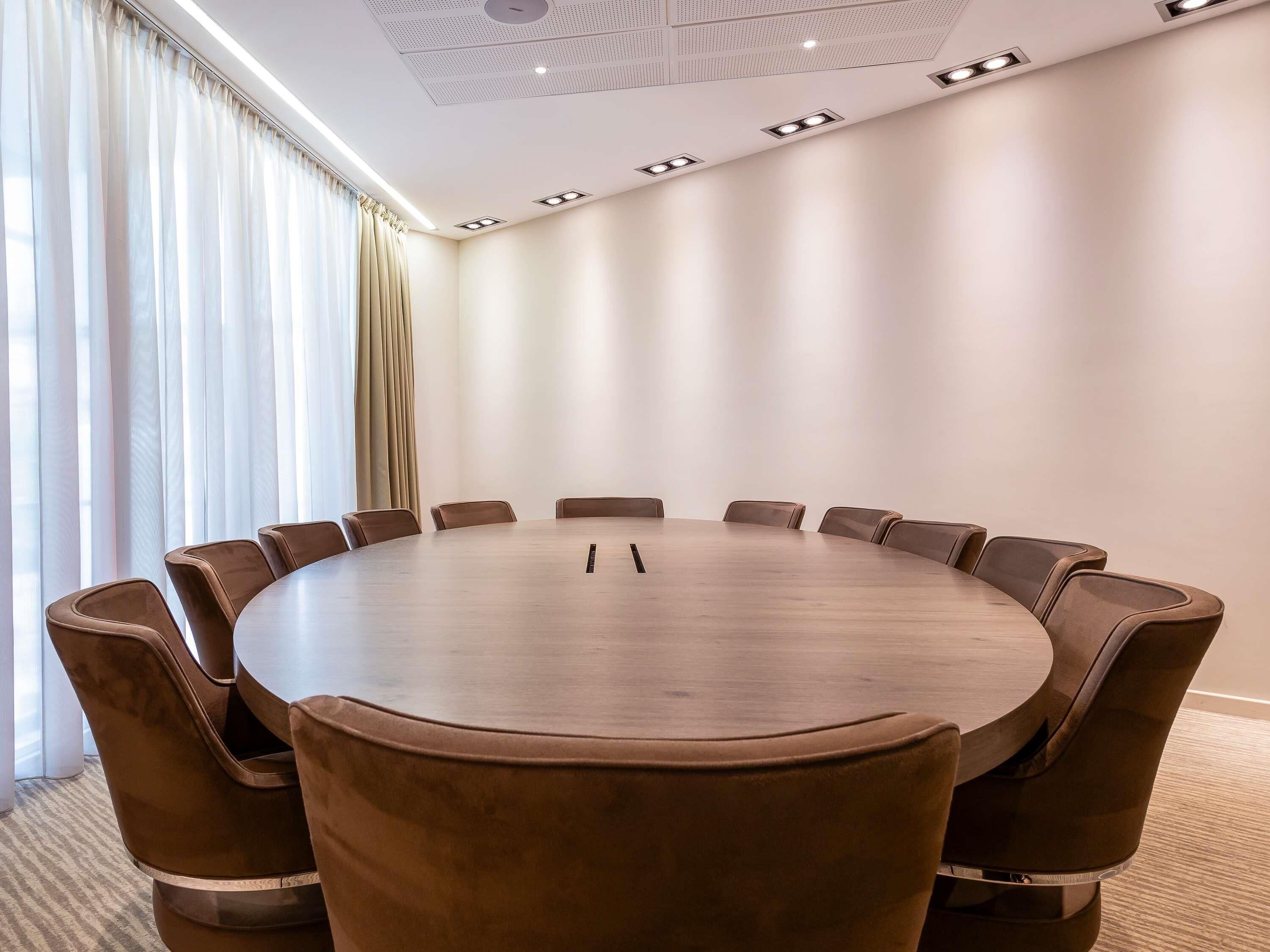 Uma sala de conferências com uma mesa redonda, cadeiras estofadas em tons de castanho e um teto com iluminação embutida.