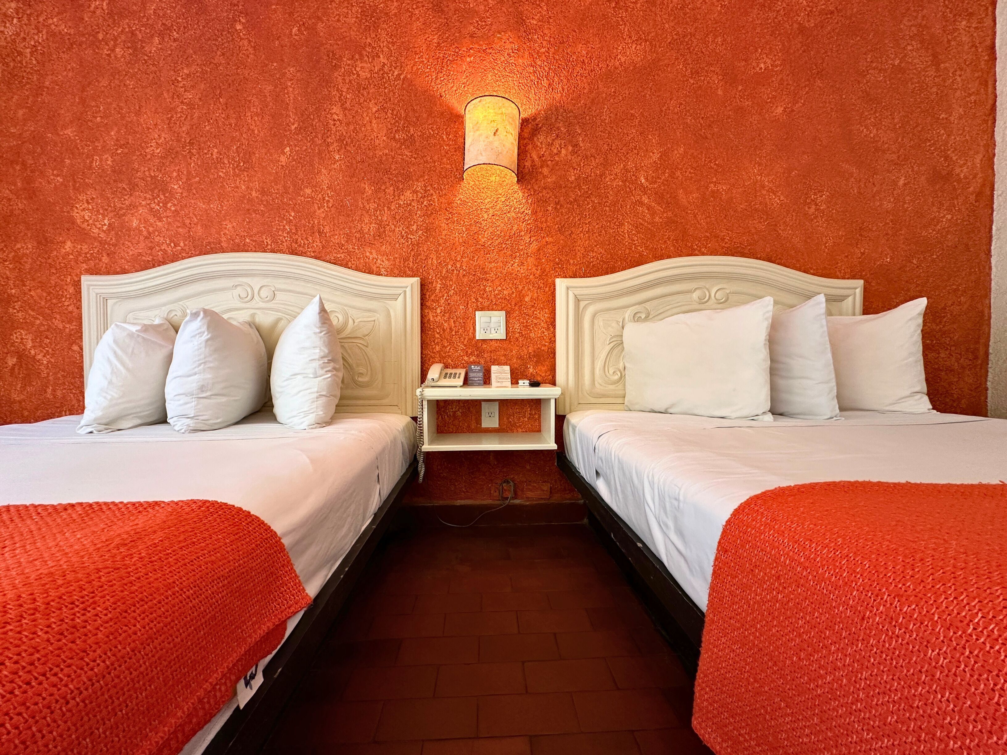 Quarto de hotel com duas camas, cada uma com roupa de cama e almofadas brancas, uma pequena mesa de cabeceira com abajur, e uma parede laranja texturizada.