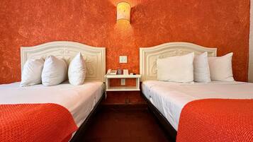 Quarto de hotel com duas camas, cada uma com roupa de cama e almofadas brancas, uma pequena mesa de cabeceira com abajur, e uma parede laranja texturizada.