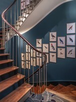 Uma escadaria com degraus de madeira e guarda-corpo metálico, levando a um patamar com ilustrações de aves emolduradas na parede.