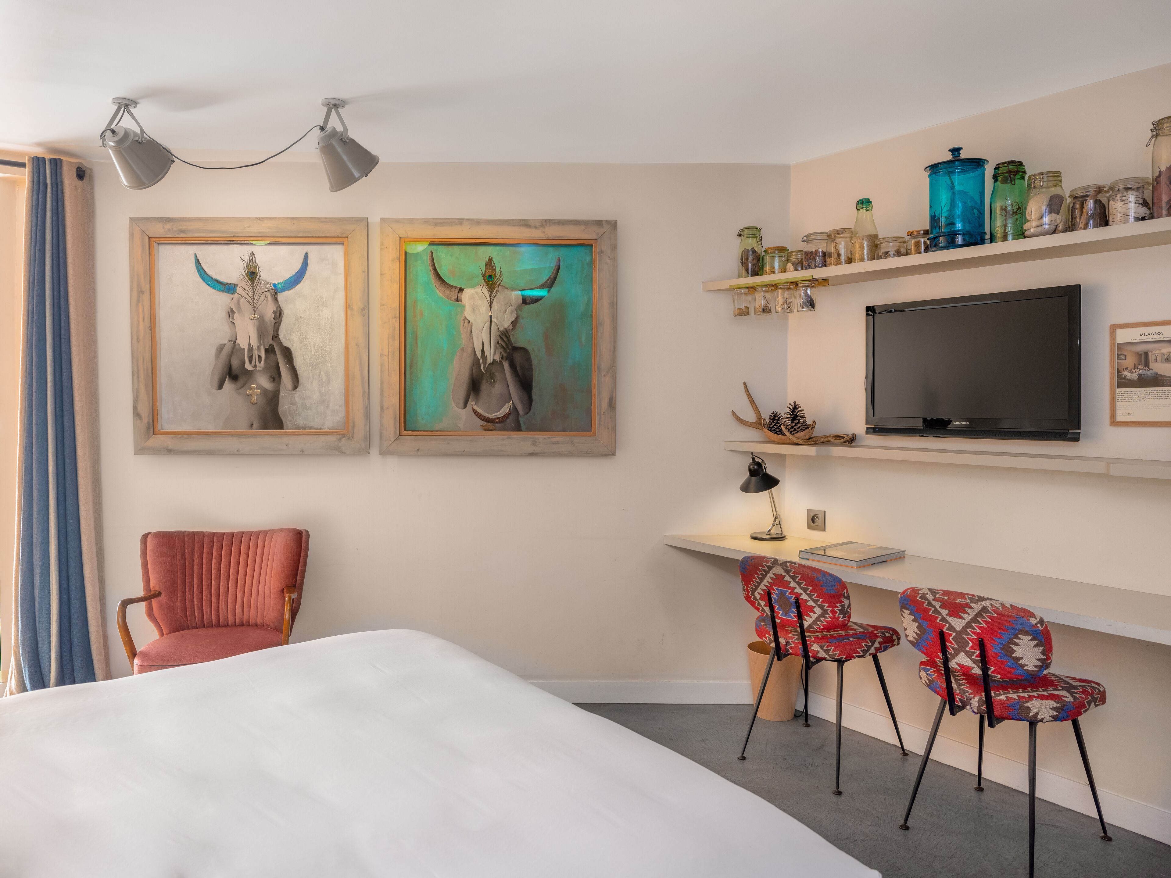 Quarto de hotel com cama, duas cadeiras, escrivaninha com computador, televisão e obras de arte na parede.
