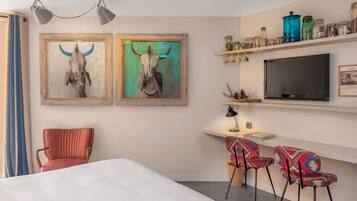 Quarto de hotel com cama, duas cadeiras, escrivaninha com computador, televisão e obras de arte na parede.
