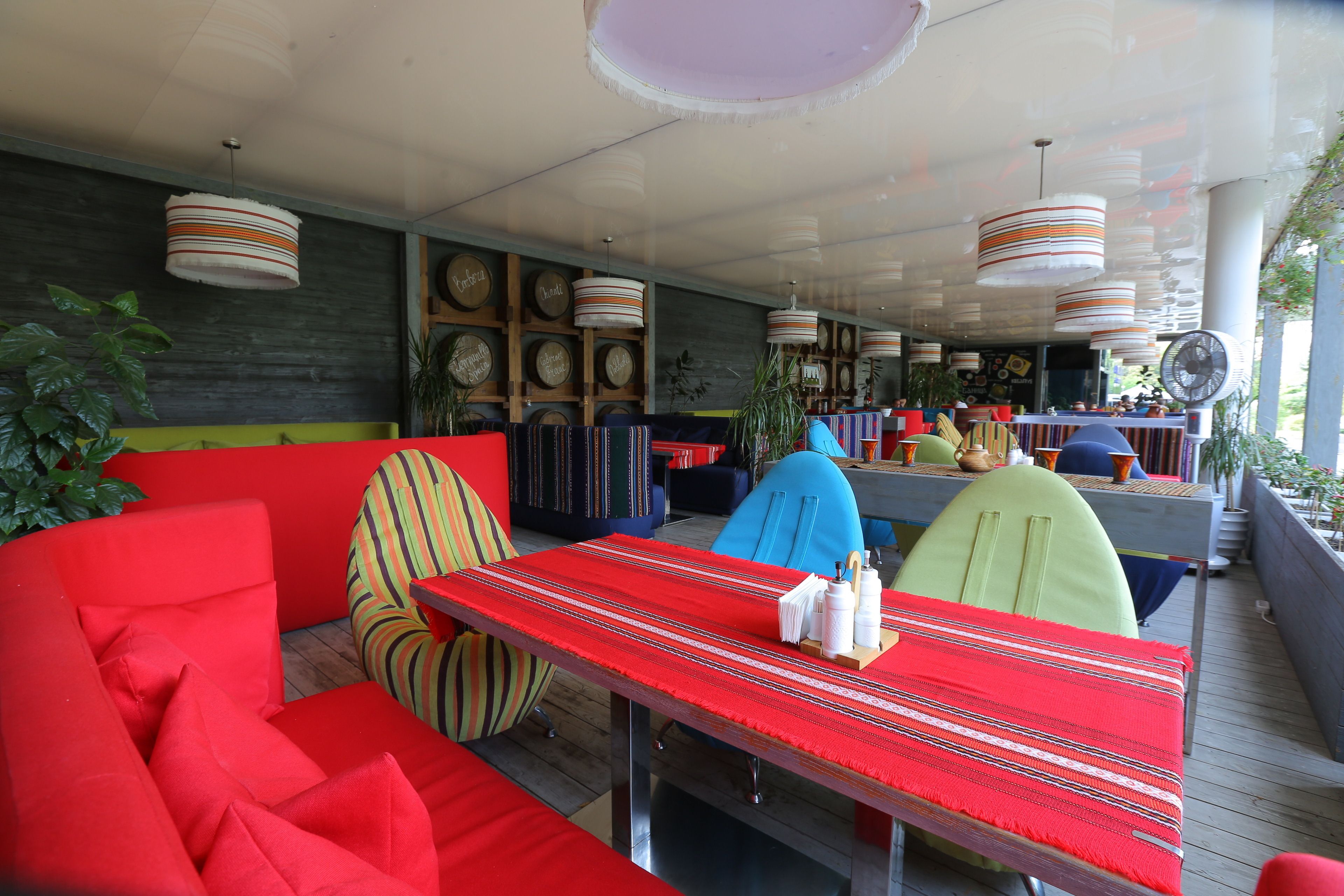 Um restaurante com assentos coloridos, uma mesa comprida com toalha vermelha e luminárias listradas penduradas.