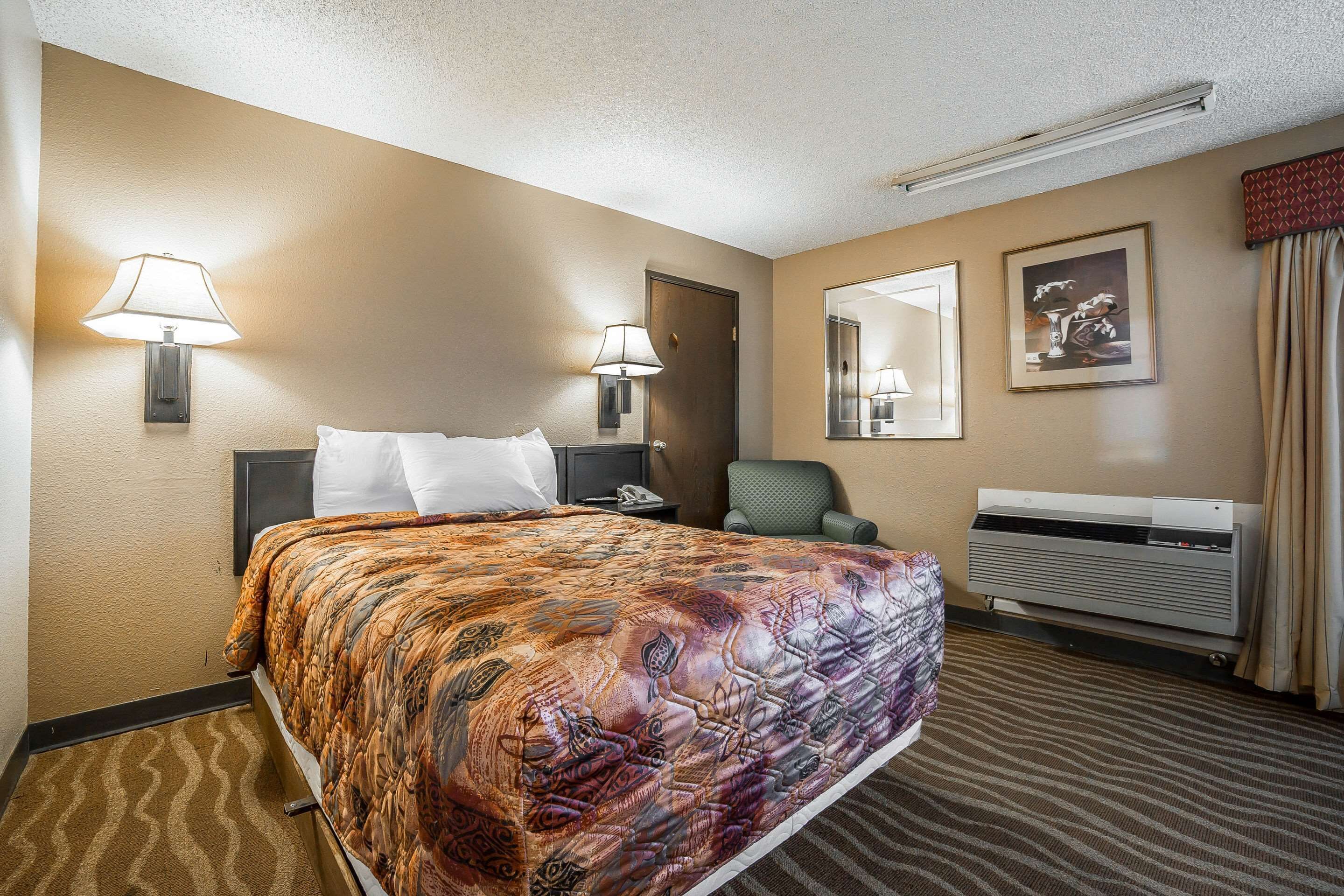 Quarto de hotel com uma cama grande, duas luminárias fixadas na parede, uma cadeira, uma escrivaninha com telefone, um espelho e um aparelho de ar condicionado.