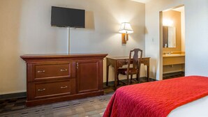 Quarto de hotel com cama, escrivaninha, cadeira, TV fixada na parede e espelho sobre o espelho do banheiro.