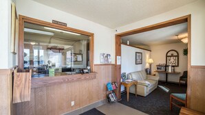 Uma área de recepção de hotel, com um balcão de madeira, uma sala de espera com sofá e poltronas, e uma kitchenette visível através do vidro.