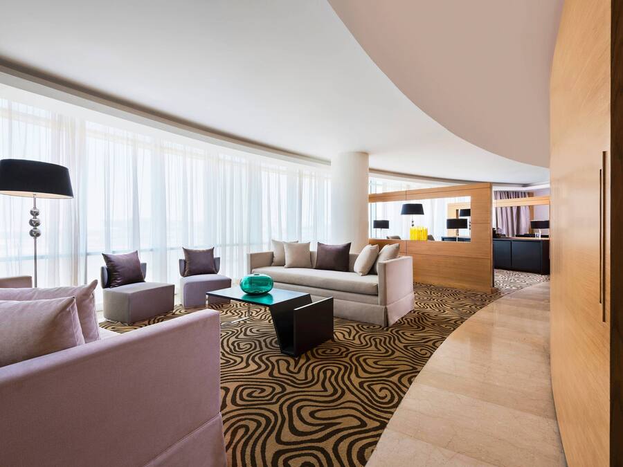 Um lobby de hotel moderno com uma parede de madeira curva, área de estar com sofás e poltronas, carpete estampado e uma grande janela com cortinas translúcidas.