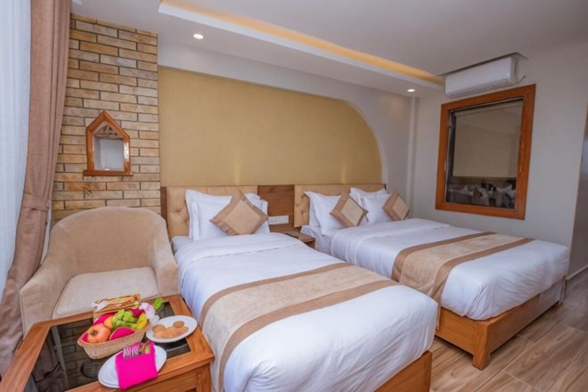 Quarto de hotel com duas camas, uma mesinha com frutas e uma cesta, uma cadeira e um espelho na parede.