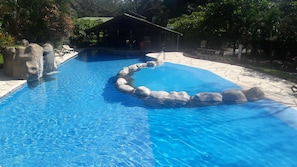 Uma piscina azul límpida com borda de pedras, área de estar coberta e um edifício ao fundo.
