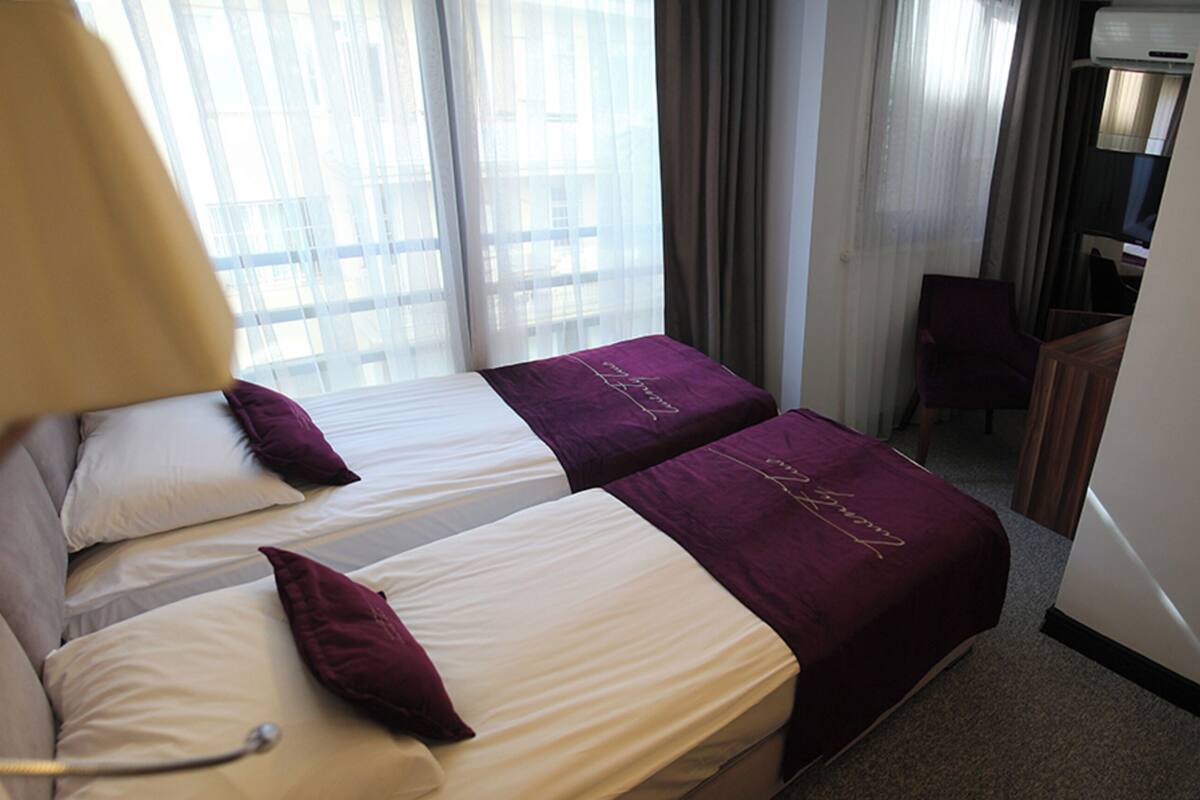 Quarto de hotel com duas camas, cada uma com colcha branca e roxa, uma mesinha de cabeceira e uma cadeira.