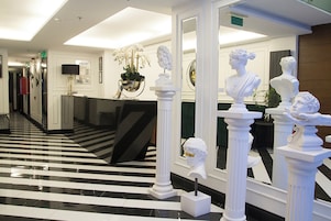 Um saguão de hotel com piso listrado em preto e branco, colunas brancas e bustos em pedestais.