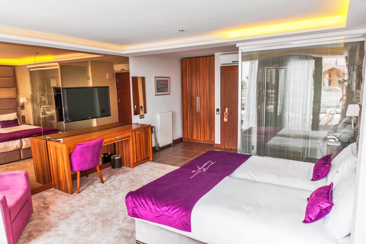 Quarto de hotel com cama, escrivaninha, cadeira, televisão e porta de vidro que se abre.
