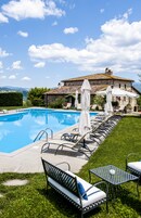 Piscina com espreguiçadeiras e guarda-sóis, um edifício de pedra ao fundo e uma área gramada.