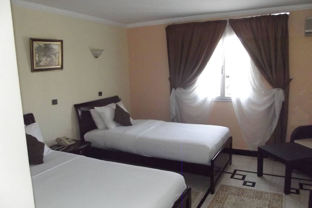 Quarto de hotel com duas camas de solteiro, uma escrivaninha, uma cadeira, uma mesa de cabeceira, uma luminária e uma janela com cortinas.