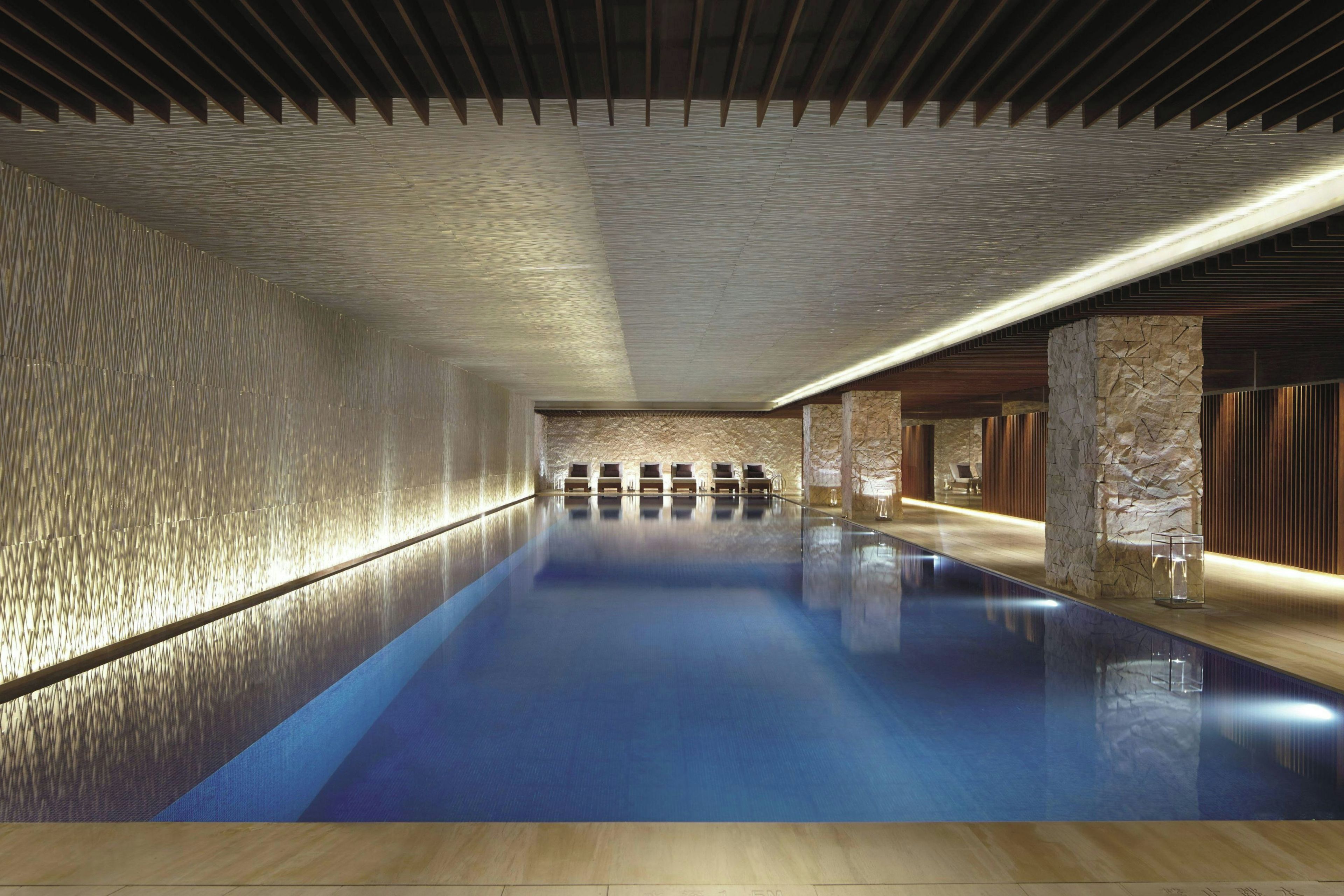 Piscina interior comprida e estreita com superfície refletora, ladeada por áreas de estar com painéis de madeira e detalhes em pedra.