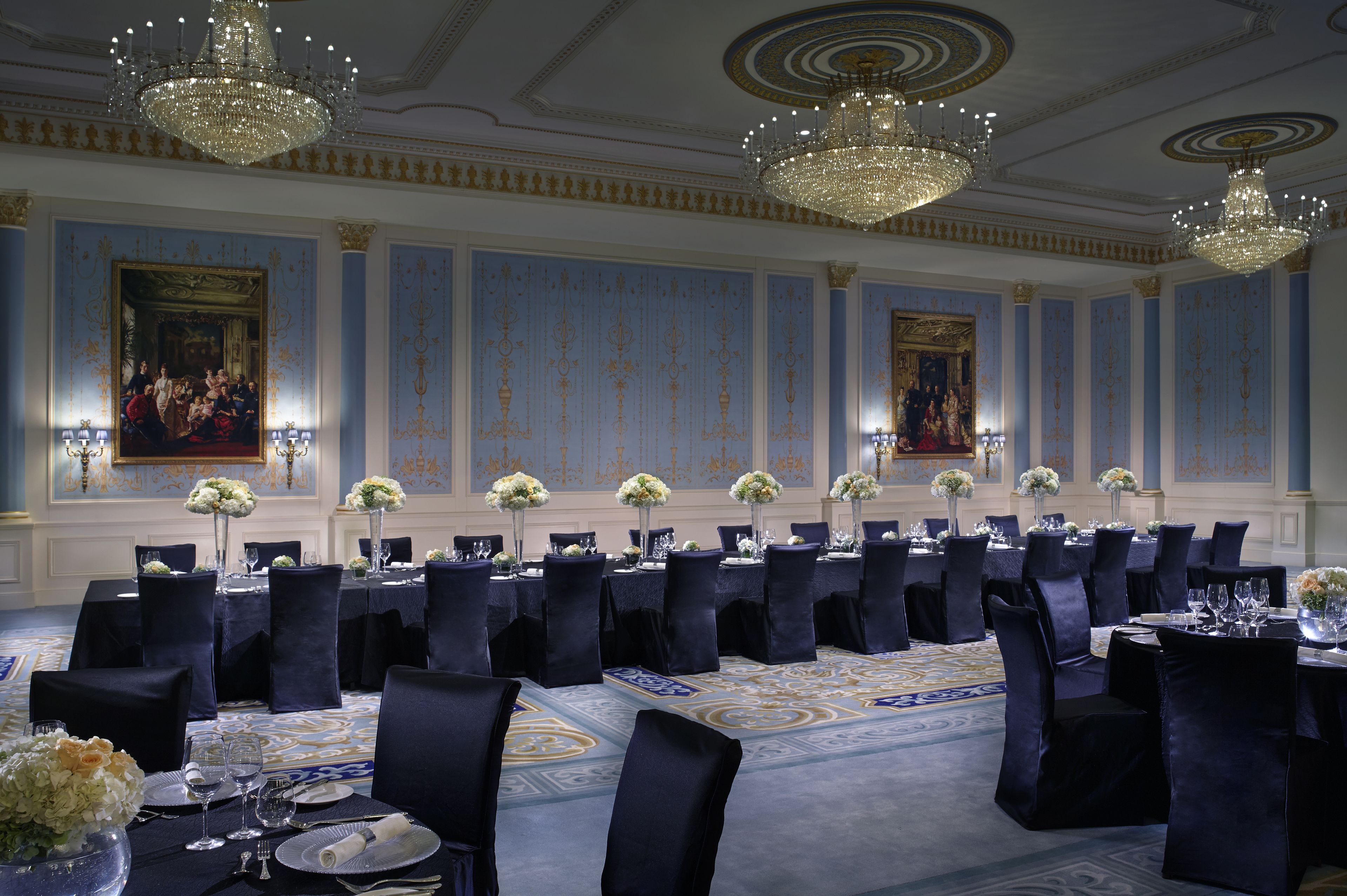Uma sala de banquetes formal, com longas mesas, cadeiras azul-escuras, lustres e pinturas clássicas nas paredes.