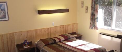 Quarto com cabeceira de madeira, mesa de cabeceira com abajur, janela com cortinas e radiador.