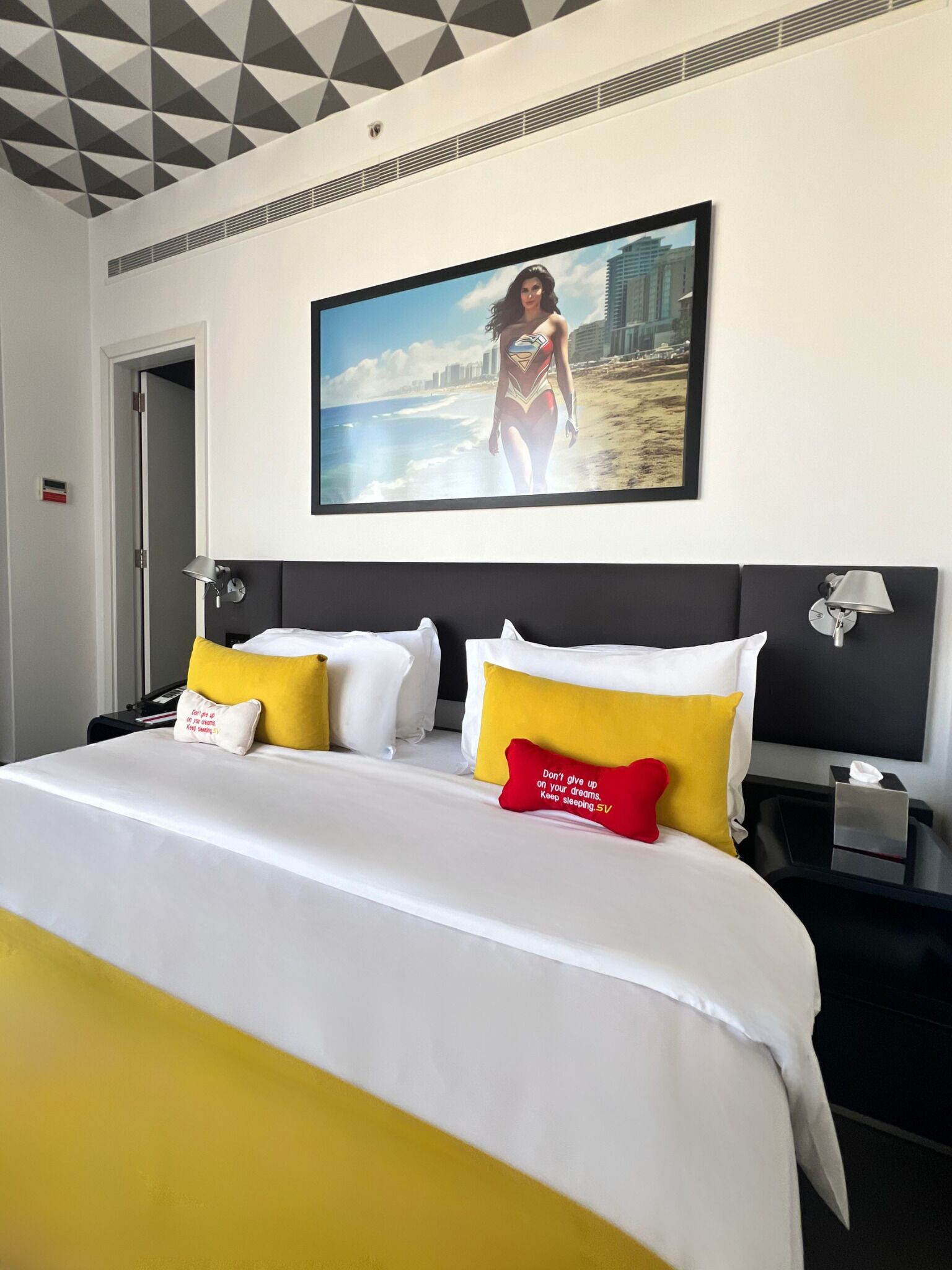 Quarto de hotel com uma cama, duas mesas de cabeceira, um criado-mudo, televisão fixada na parede e um design geométrico no teto.