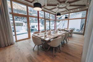 Uma sala de jantar com mesa de madeira posta para seis pessoas, grandes janelas com vista para uma montanha nevada e piso de madeira.