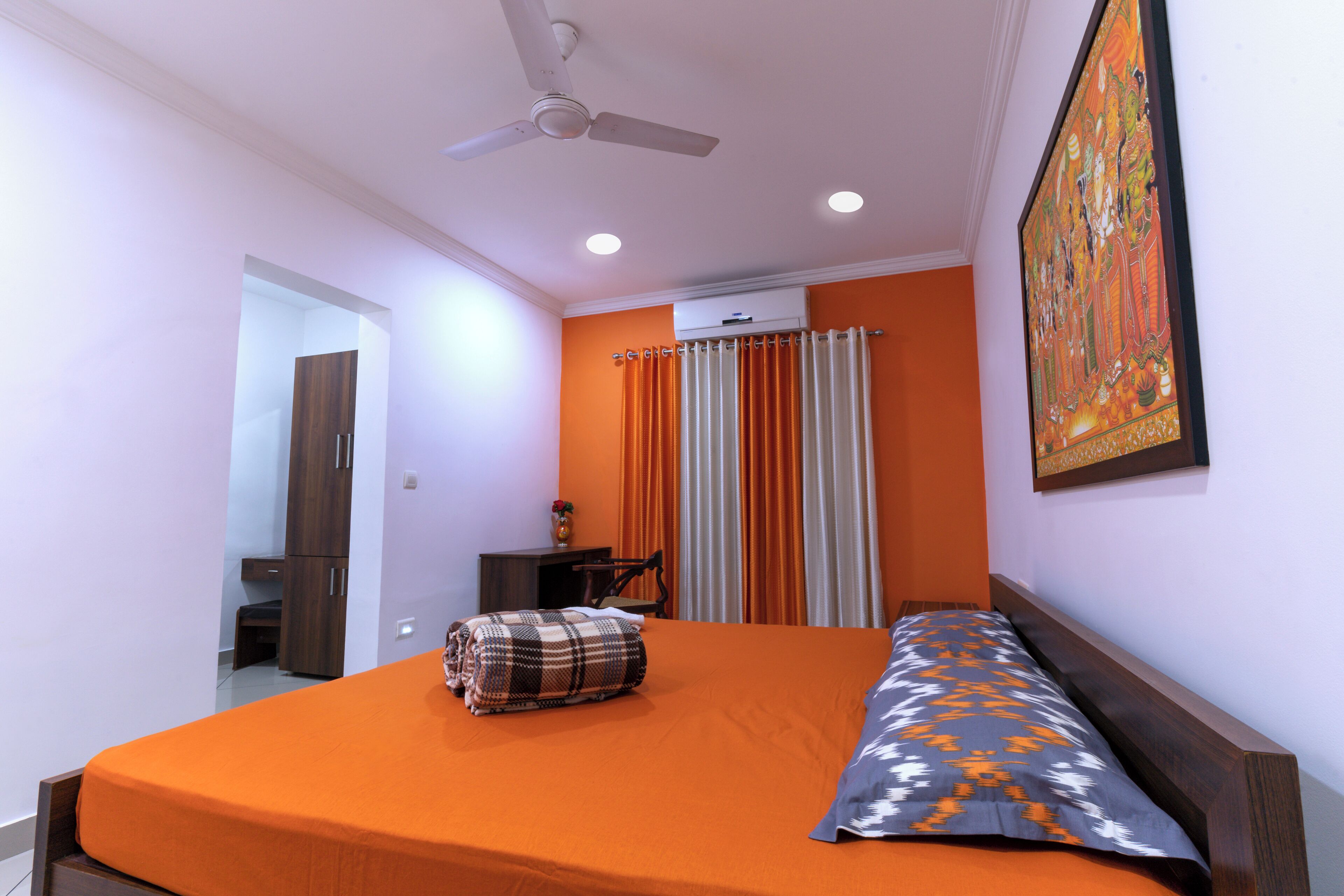 Um quarto com colcha laranja, ventoinha no teto, janela com cortinas laranjas e um quadro pendurado na parede.