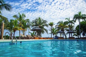 Piscina com espreguiçadeiras e guarda-sóis, rodeada por palmeiras.