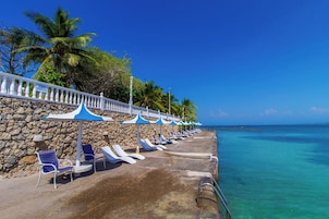 Um resort à beira-mar com uma fileira de espreguiçadeiras e guarda-sóis, uma parede de pedra e vista clara para o oceano.