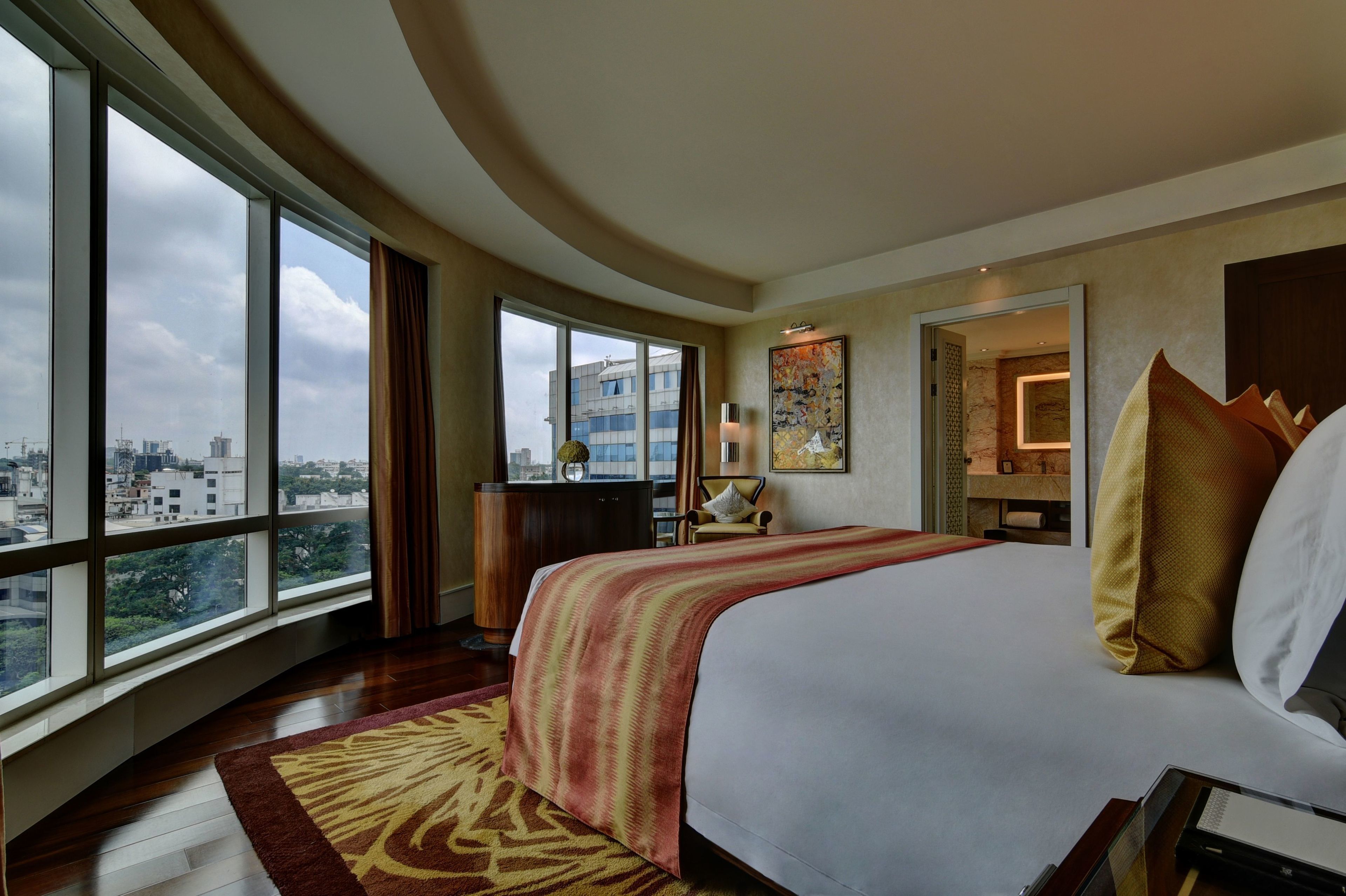 Quarto de hotel com uma cama grande, uma escrivaninha e vista da cidade através de amplas janelas.