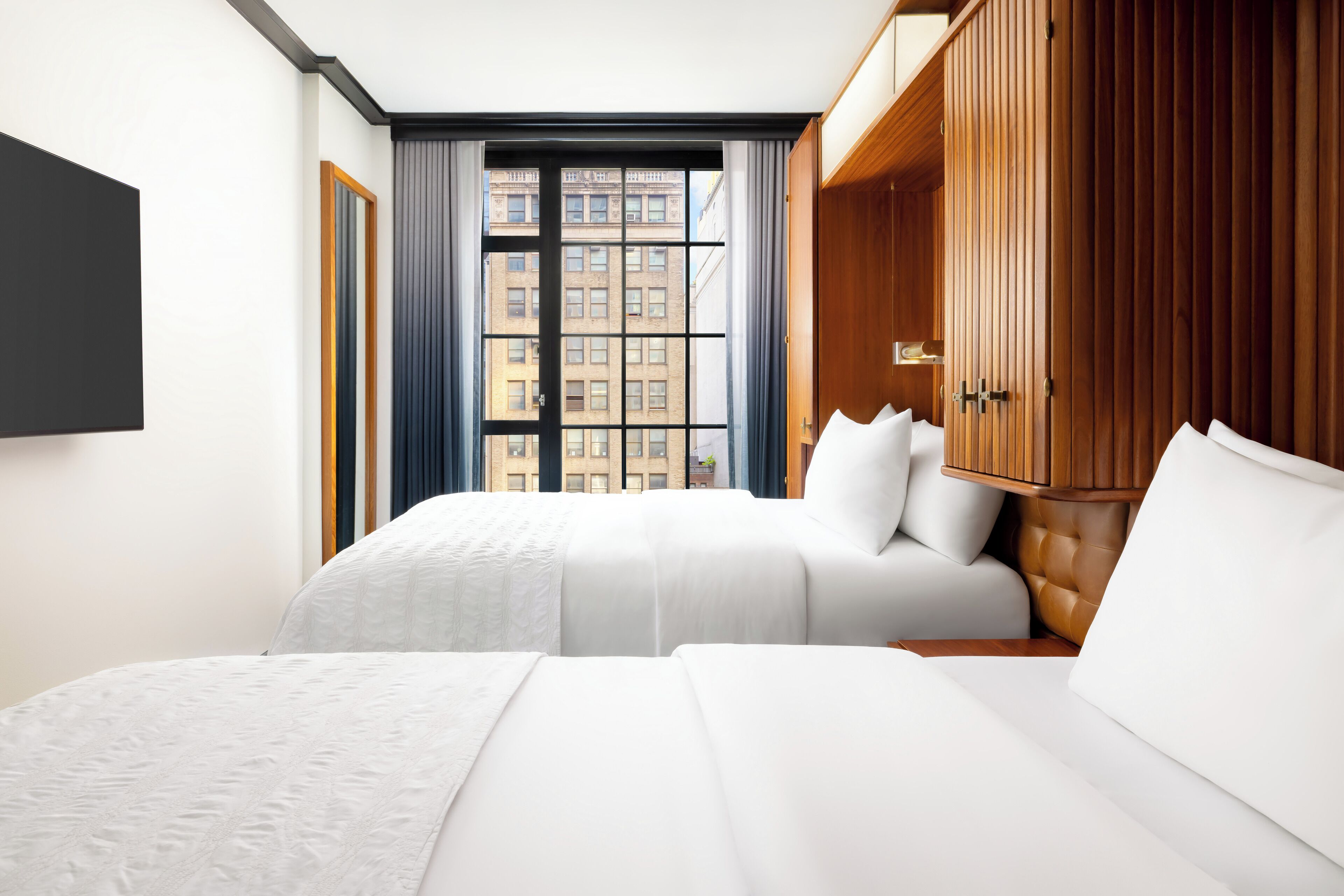 Quarto de hotel com duas camas, uma janela ampla com vista para a cidade, guarda-roupa de madeira e televisão de tela plana na parede.