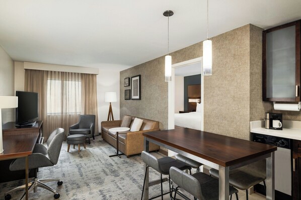 Quarto de hotel com escrivaninha de madeira, duas poltronas, um sofá, televisão, candeeiro, máquina de café e espelho.