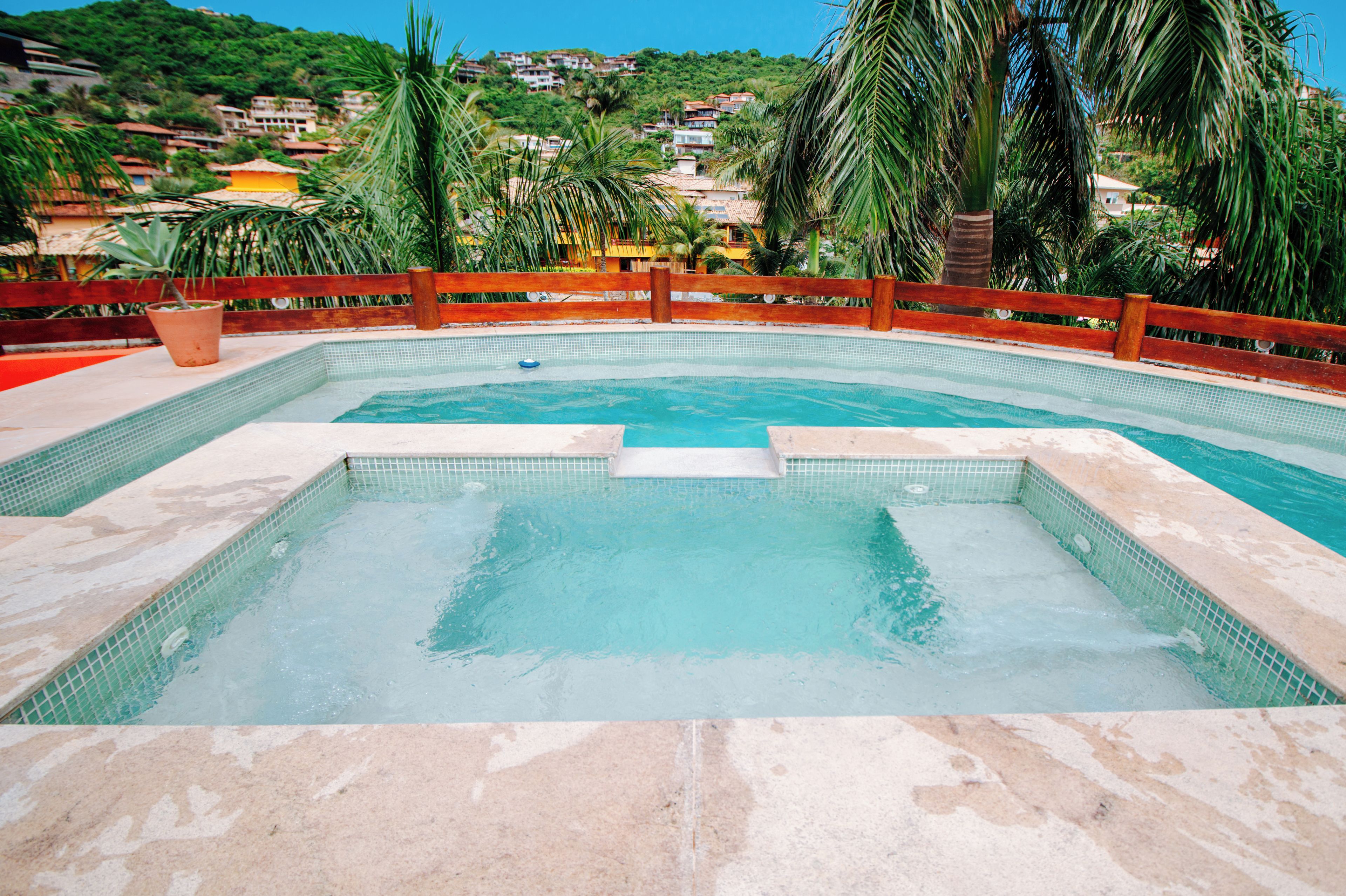Piscina retangular com borda azulejada, cercada por uma vedação de madeira e palmeiras ao fundo.