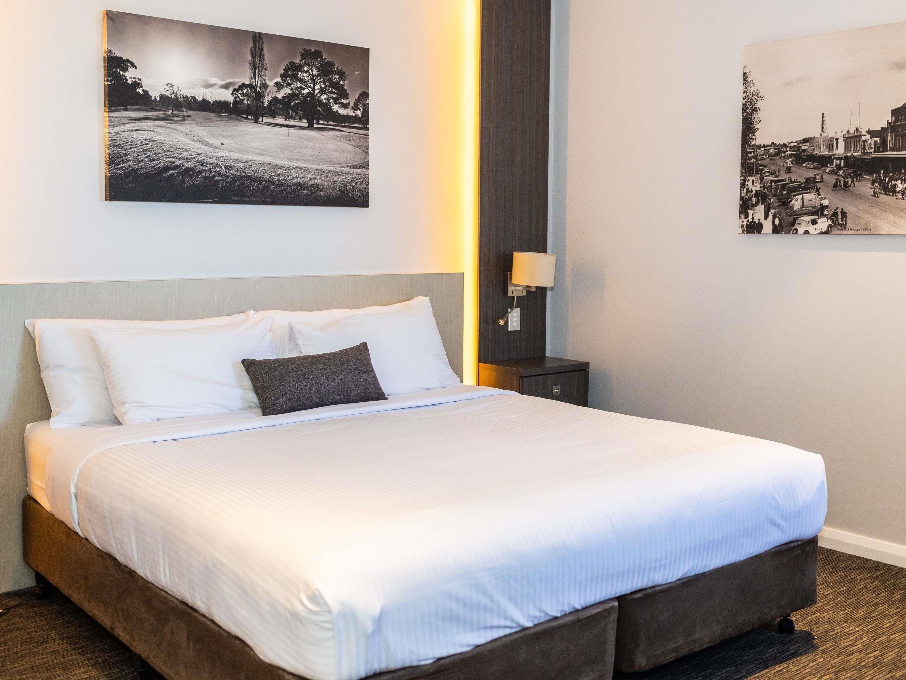 Quarto de hotel com uma cama, mesa de cabeceira, abajur e duas imagens penduradas na parede.