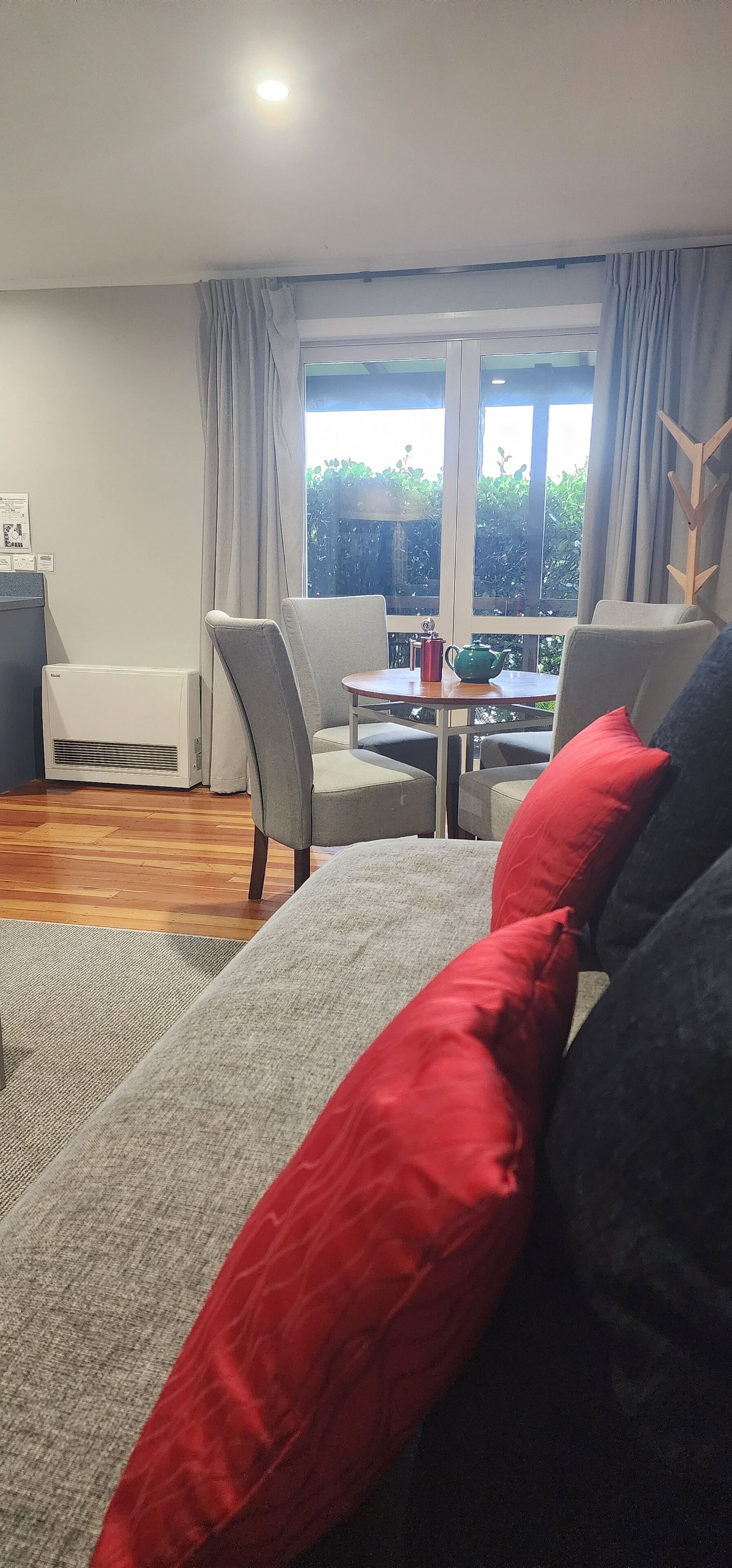 Uma sala de estar moderna com um sofá cinzento, almofadas vermelhas e pretas, uma mesa pequena com uma chaleira vermelha e vista para o verde através de janelas amplas.