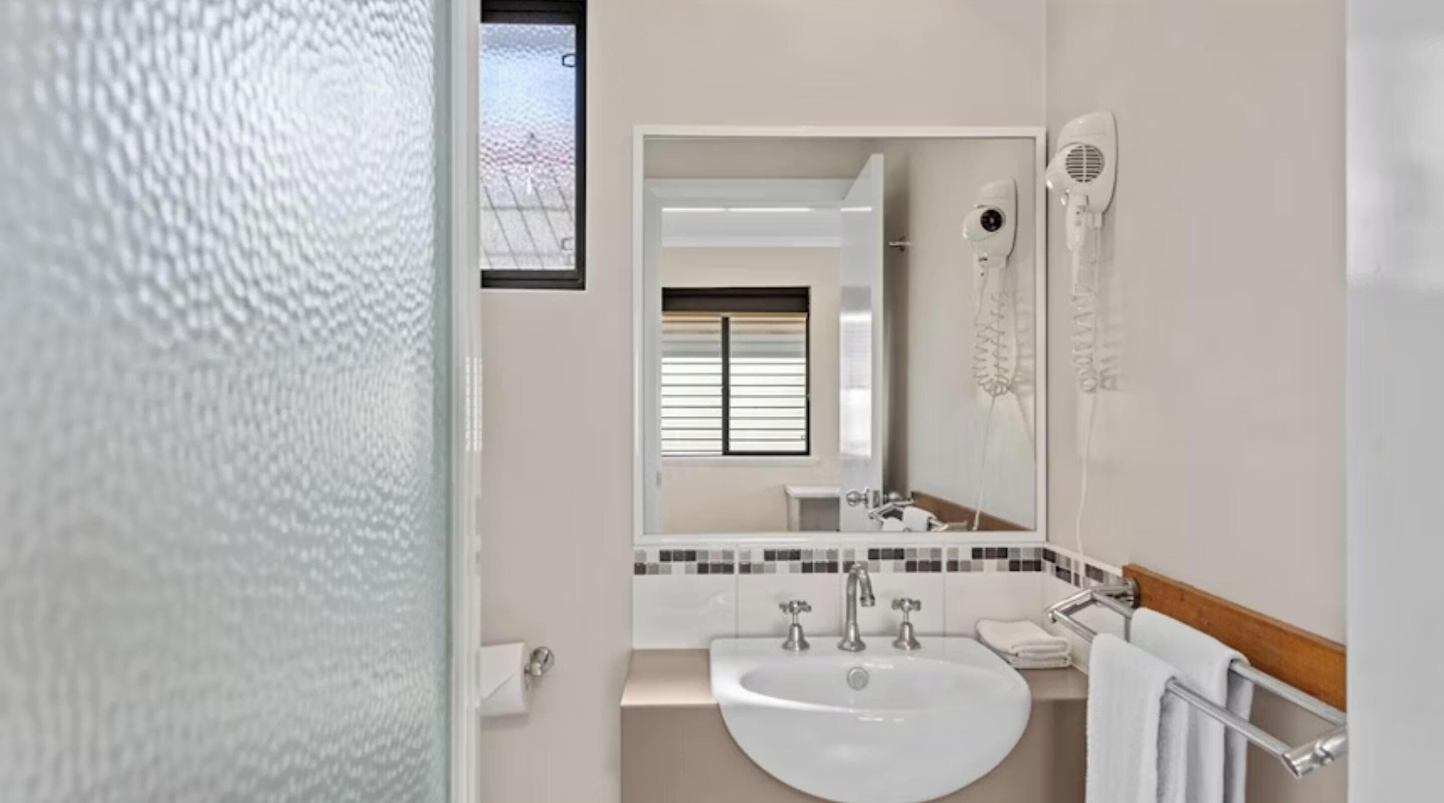 Uma casa de banho com uma pia branca, duas torneiras, um espelho e dois secadores de cabelo fixados na parede.