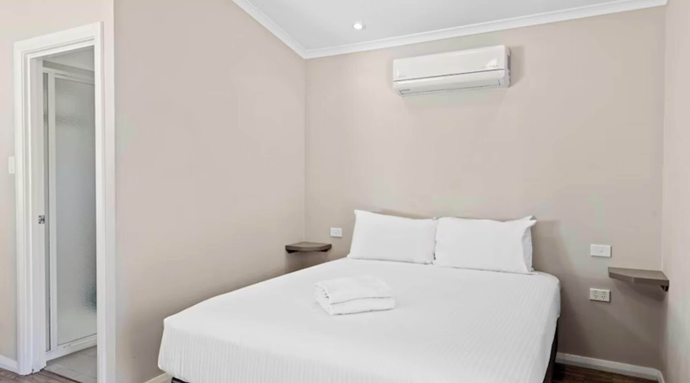 Quarto com cama, roupa de cama branca, mesa de cabeceira, ar condicionado fixado na parede e uma porta.
