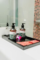 Uma bancada de banheiro com duas garrafas de shampoo e condicionador SleepWithin, uma flor rosa e um porta-objetos com toalhas vermelhas.