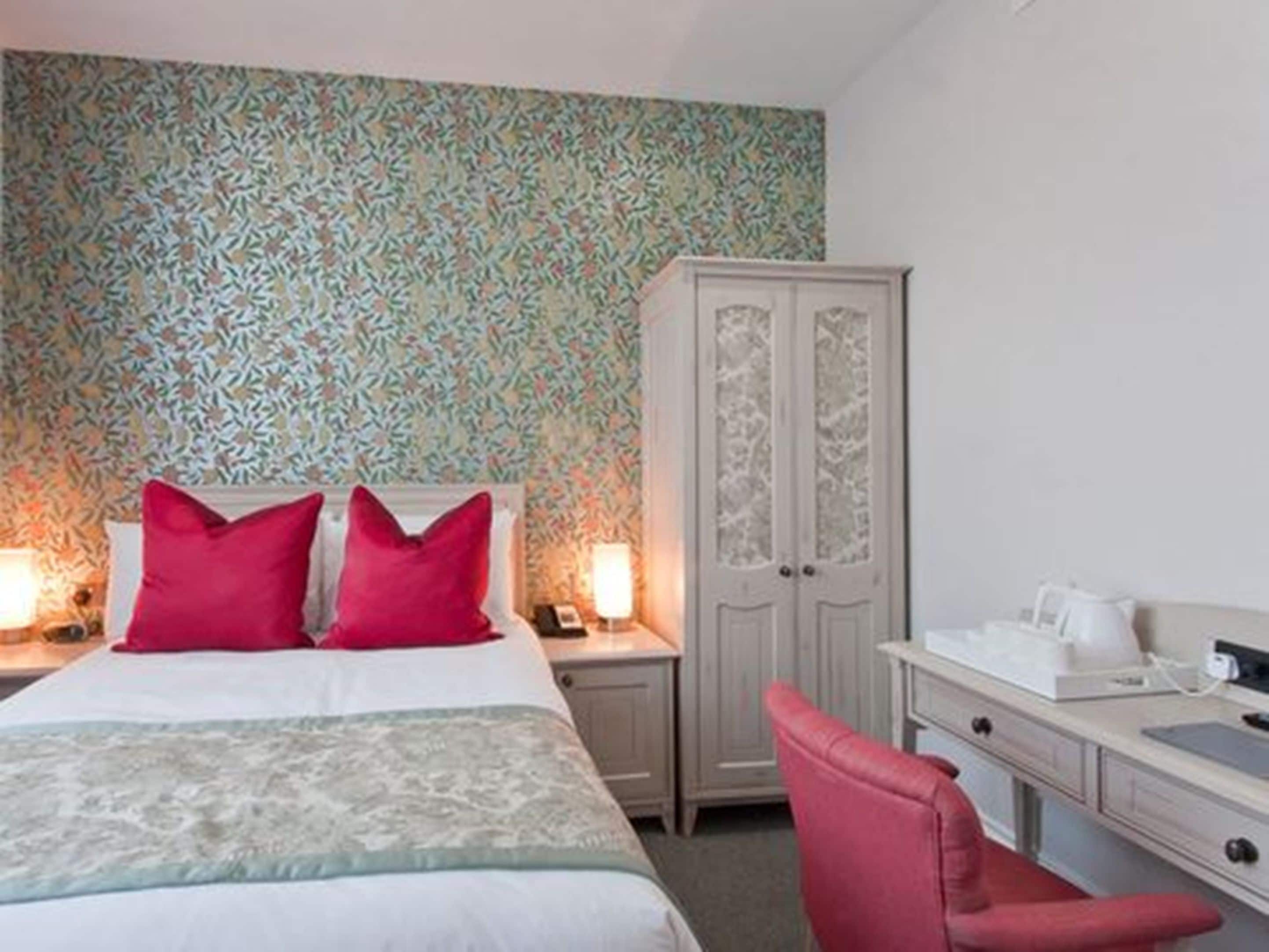 Quarto de hotel com uma cama, duas almofadas vermelhas, uma mesa de cabeceira com abajur, um guarda-roupa, uma escrivaninha com cadeira e uma cafeteira.