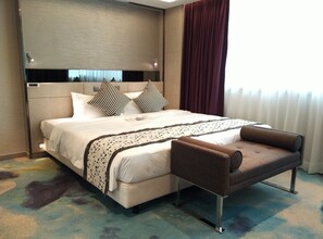 Quarto de hotel com uma cama grande, uma poltrona marrom, cabeceira com painel claro e uma janela com cortina escura.